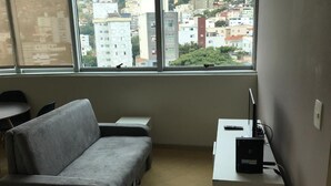 Quarto pequeno e moderno com sofá cinza, uma mesinha e uma TV em um suporte. Uma janela grande oferece vista para paisagem urbana com edifícios e áreas verdes.