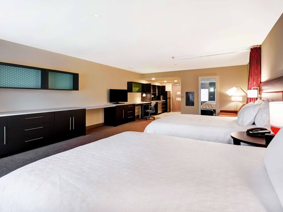 Quarto de hotel com duas camas, uma escrivaninha, televisão e cozinha compacta.