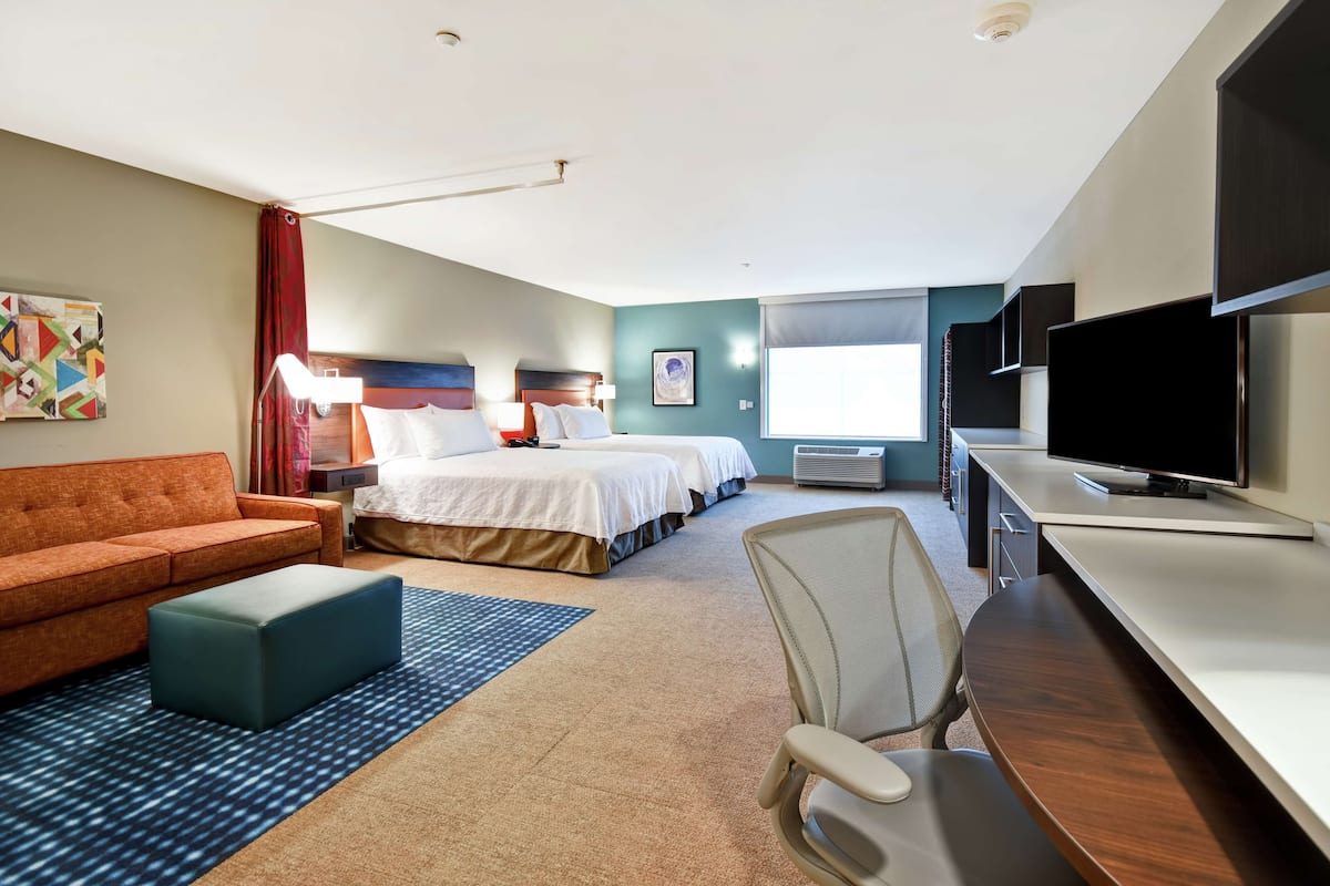 Quarto de hotel com sofá, duas camas, escrivaninha com televisão e uma janela grande.