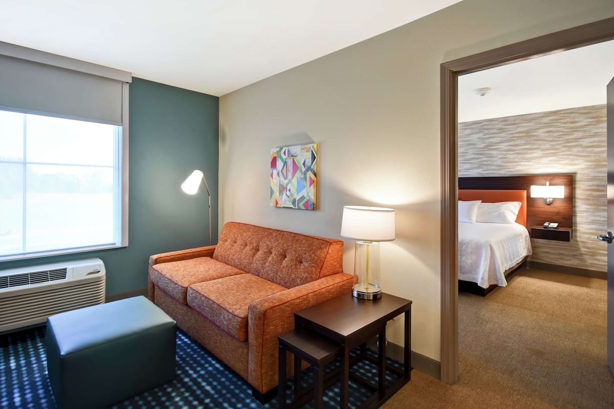 Quarto de hotel com sofá, uma mesinha com abajur, cama com cabeceira e um quadro na parede.