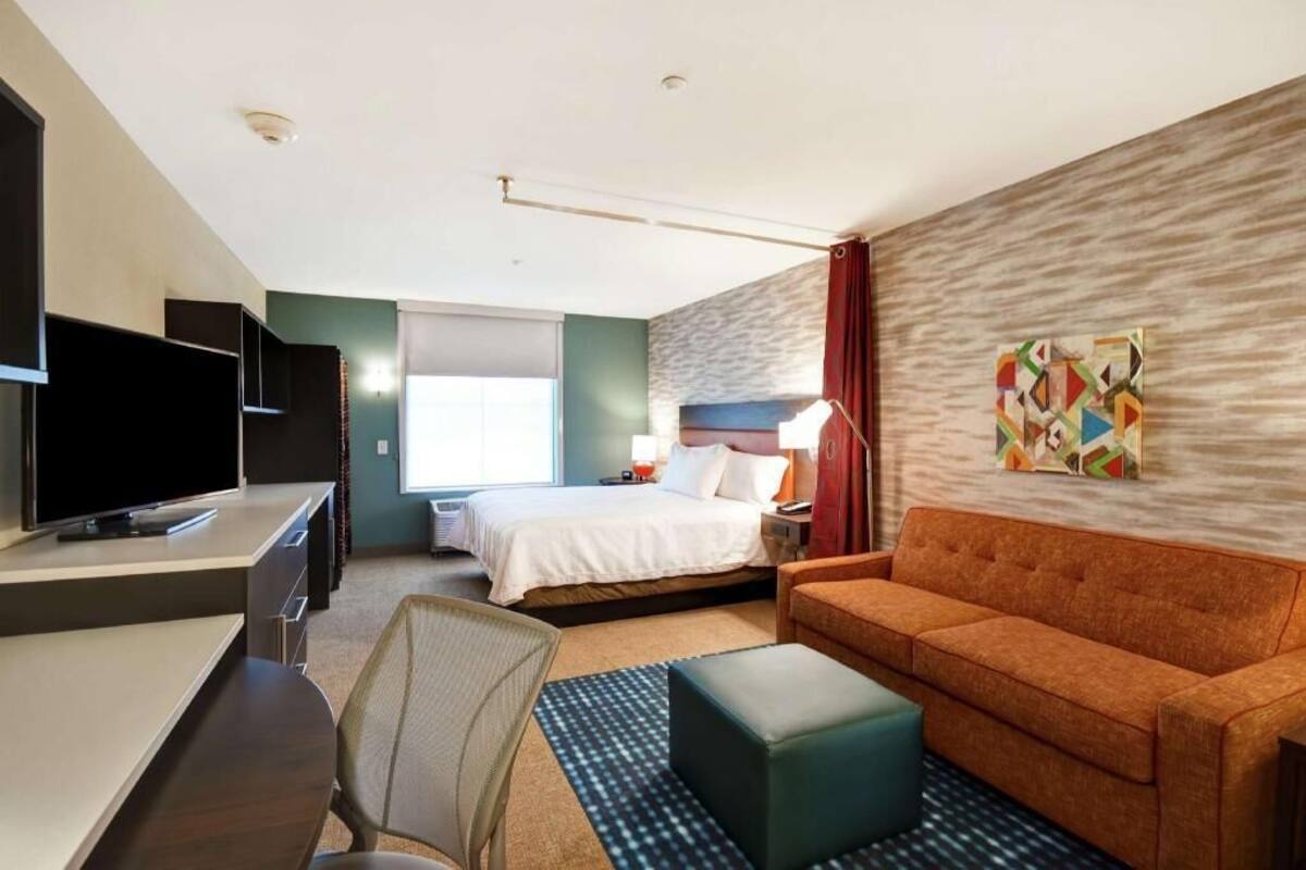 Quarto de hotel com uma cama, uma mesa com televisão de tela plana, um sofá, uma poltrona e uma obra de arte na parede.