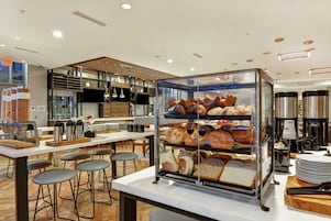 Café moderno com estação de café da manhã por autoatendimento, área de estar e vitrine com doces.