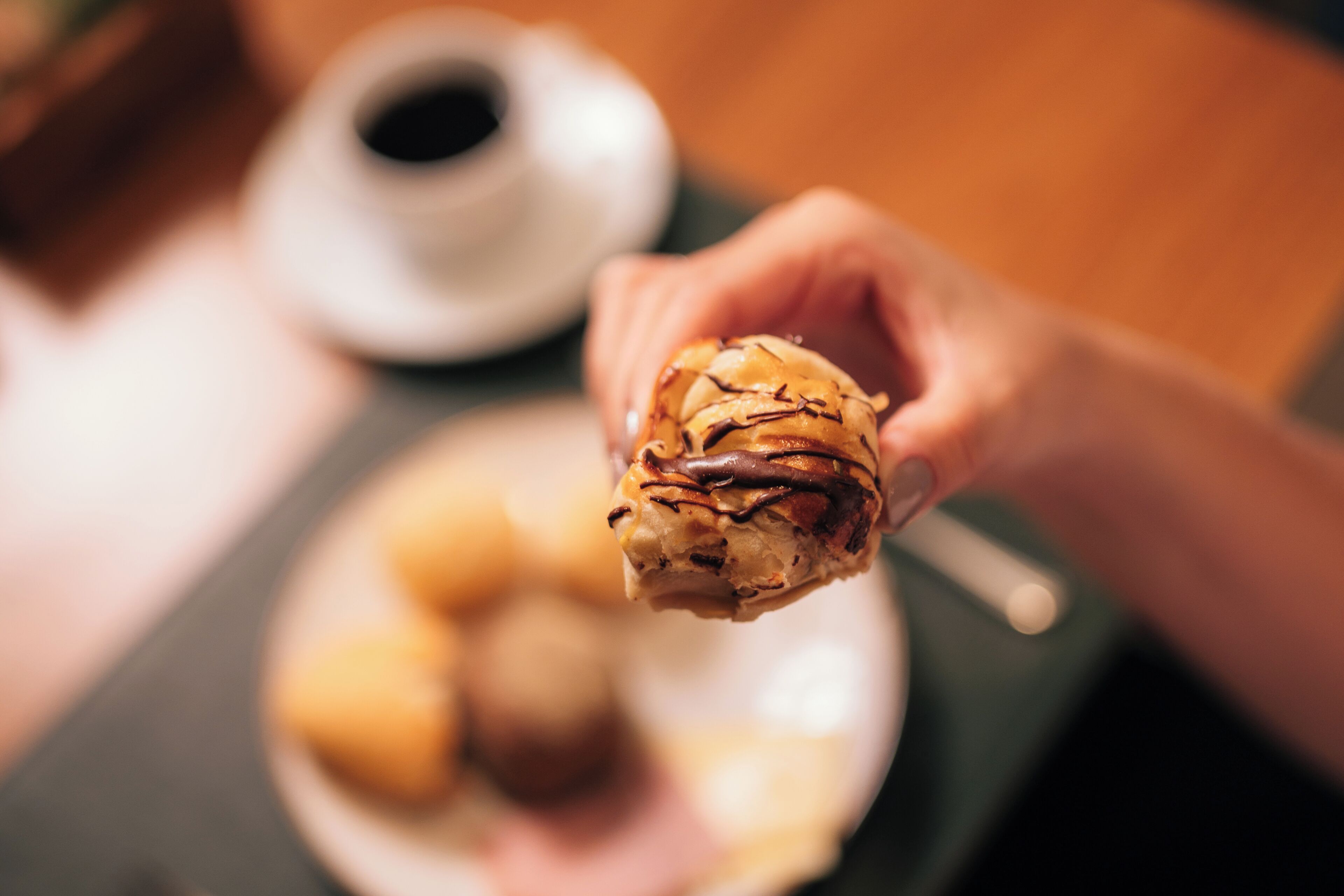 Uma mão segurando um doce com calda de chocolate, uma xícara de café e um prato com outros doces ao fundo.