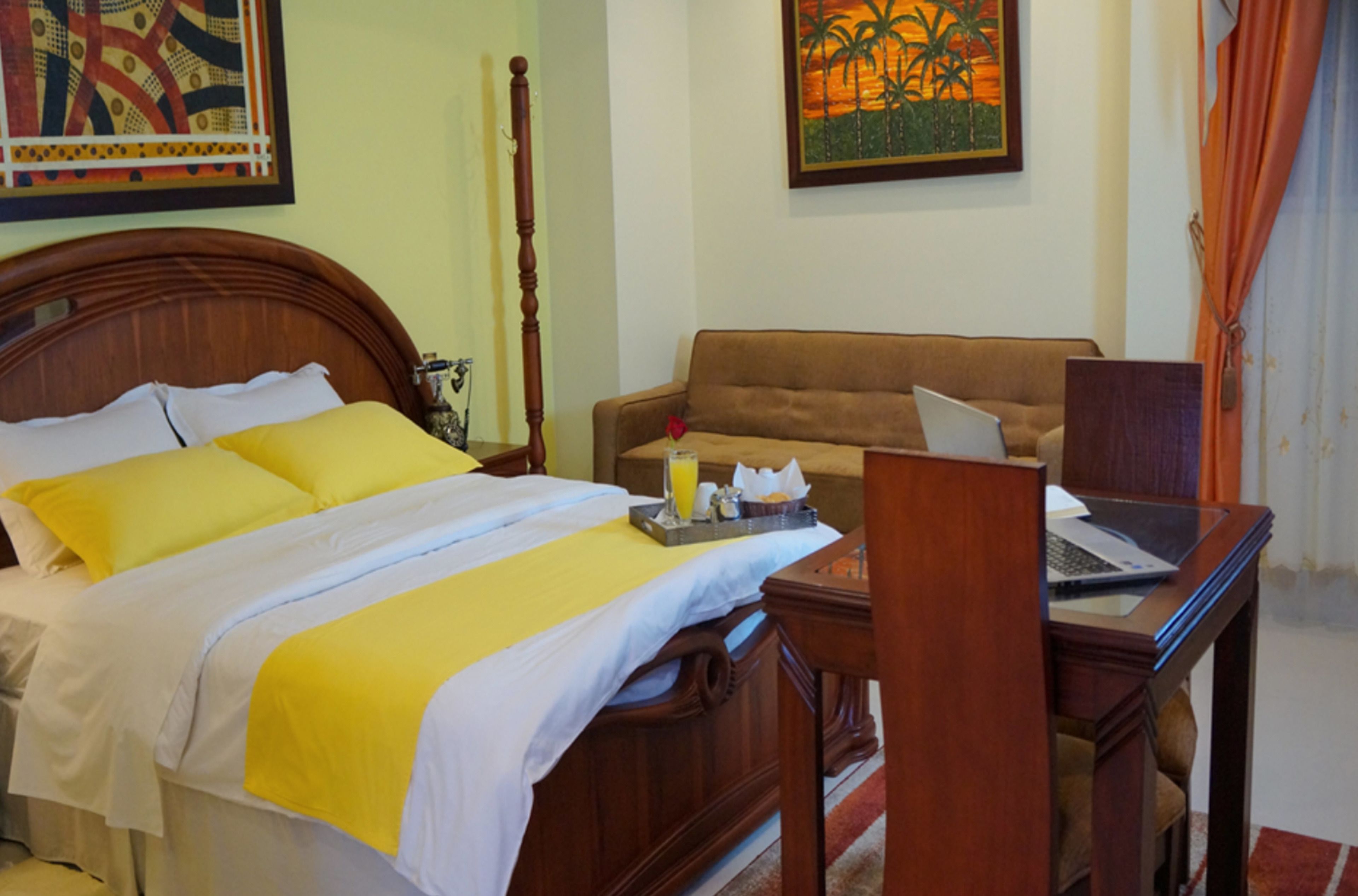 Quarto de hotel com cama de madeira, um sofá, uma escrivaninha com um laptop e um quadro na parede.