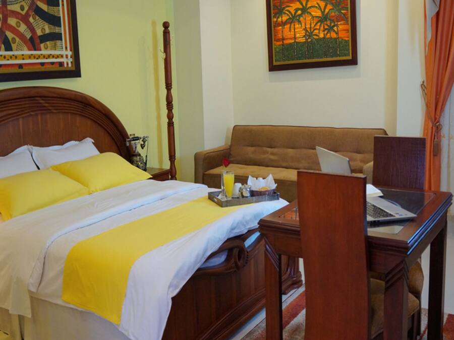 Quarto de hotel com cama de madeira, um sofá, uma escrivaninha com um laptop e um quadro na parede.