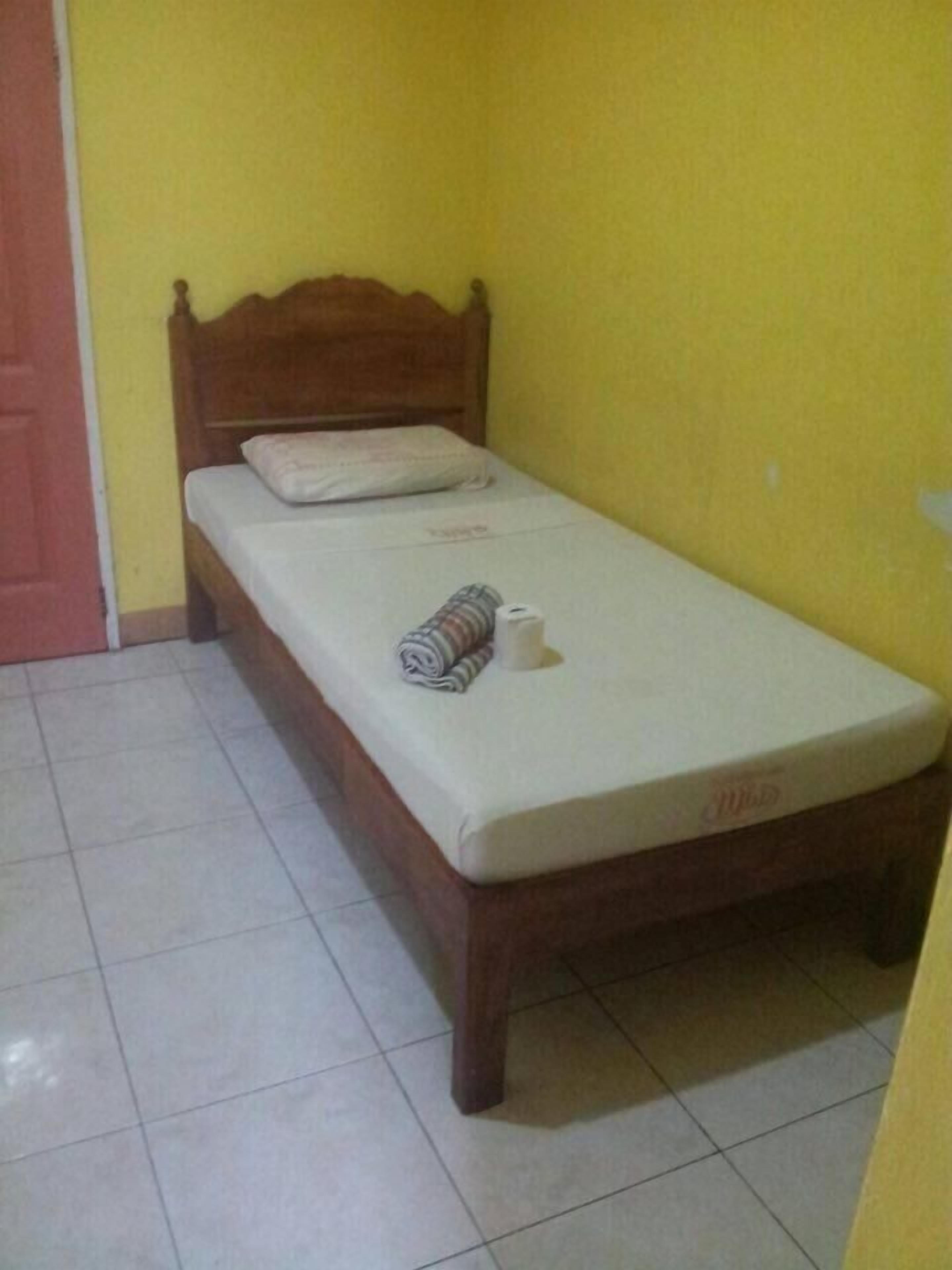 Uma cama de solteiro com cabeceira de madeira num quarto com paredes amarelas e soalho de azulejos brancos.