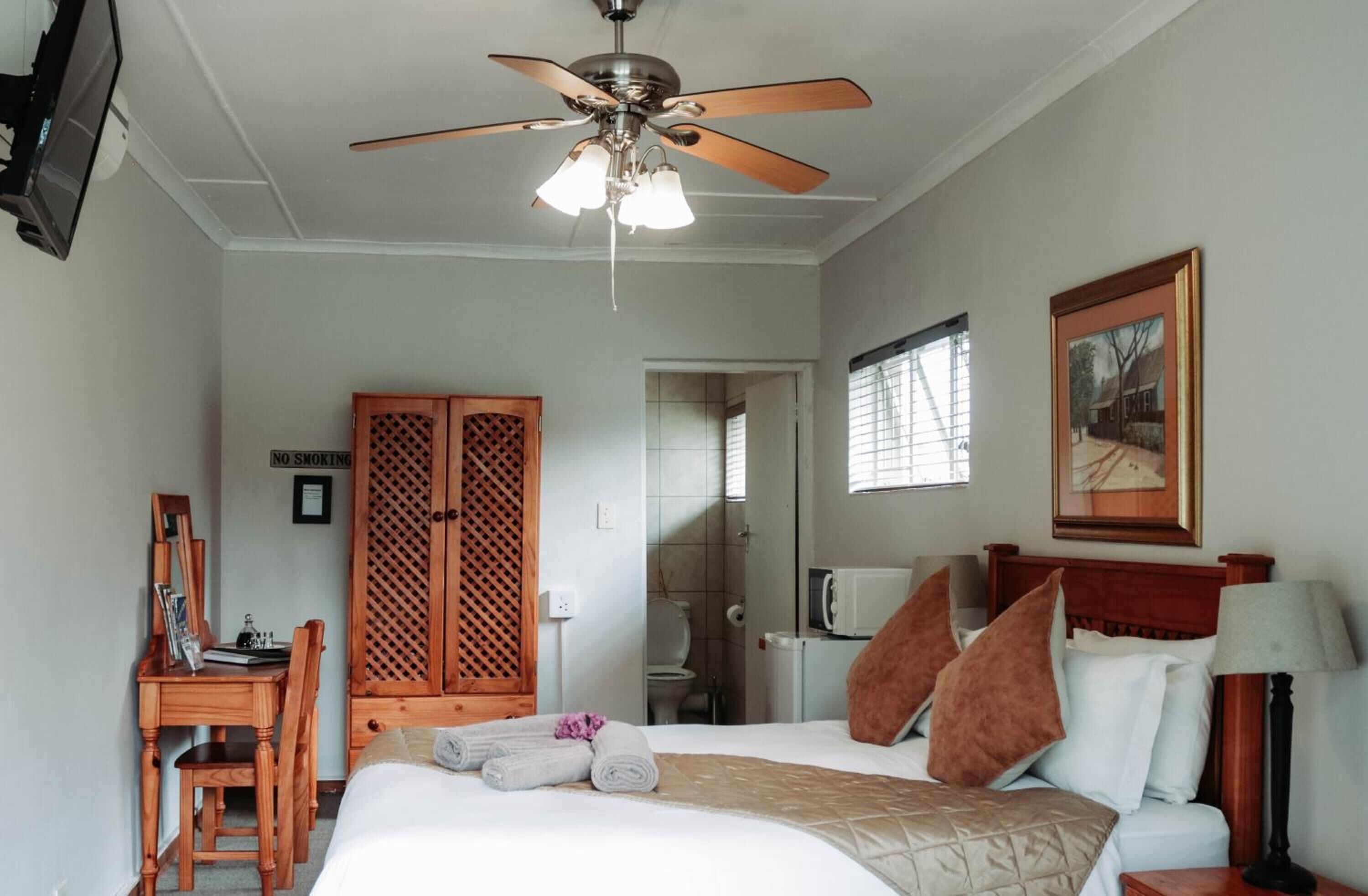 Quarto de hotel com cama, escrivaninha, cadeira, ventoinha no teto, televisão e área de chuveiro.