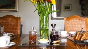 Uma mesa de jantar com um vaso de flores amarelas, um pote de vidro com geleia, um pote de vidro com mel e uma bandeja de sanduíches.