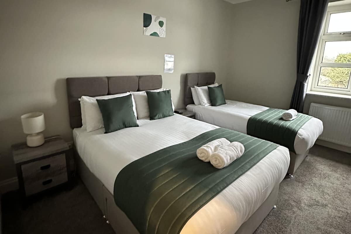 Quarto de hotel com duas camas, uma mesinha de cabeceira, uma luminária e uma janela com cortinas.