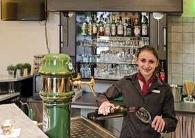 Uma área de bar com um bartender preparando uma bebida, um coqueteleira verde e prateleiras repletas de garrafas e copos.