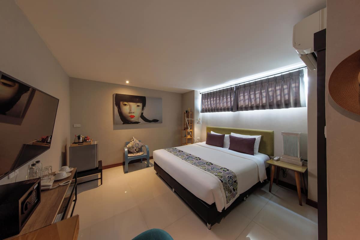 Quarto de hotel moderno com uma cama grande, uma mesa de escritório com um computador, uma cadeira e uma pequena mesa com abajur.