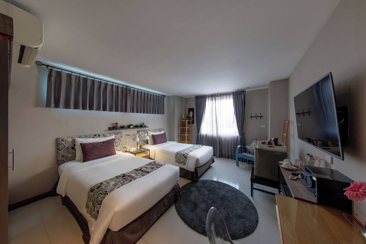 Quarto de hotel com duas camas, uma escrivaninha, uma cadeira, uma televisão e uma janela com cortinas.