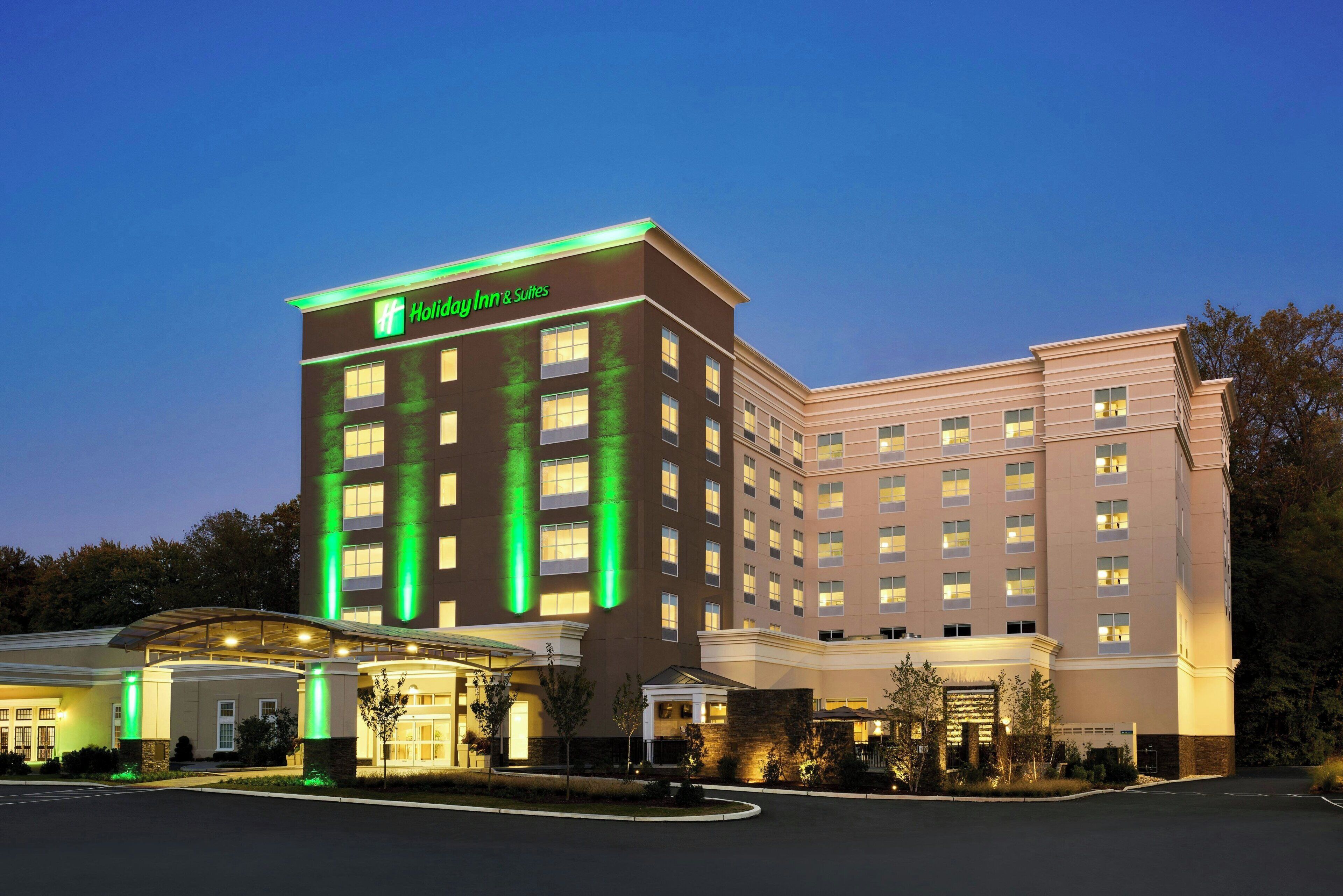 Um hotel Holiday Inn & Suites com design moderno, destacando uma placa verde chamativa, vários andares e uma entrada bem iluminada.
