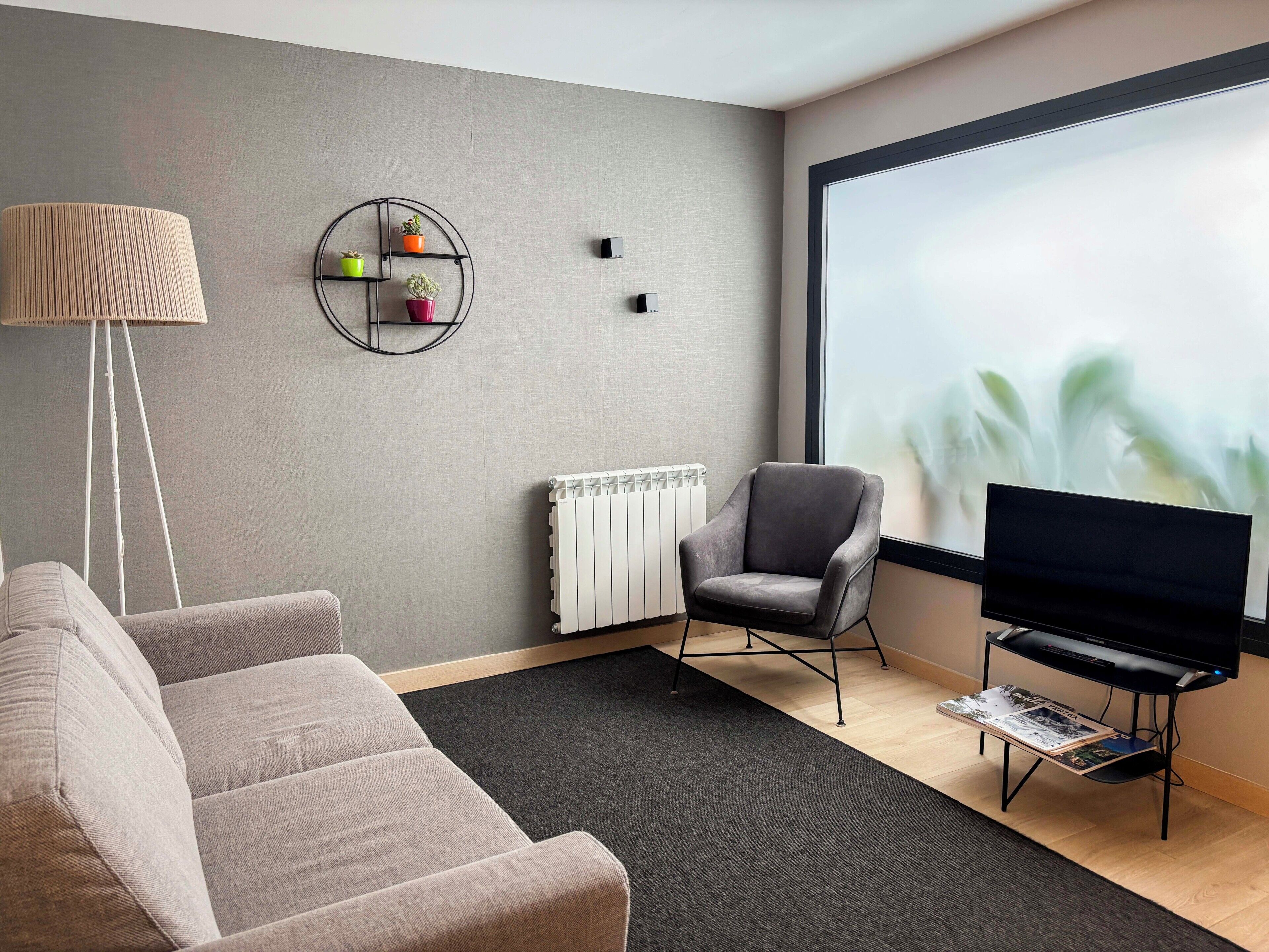 Uma sala de estar moderna com um sofá cinzento, uma poltrona cinzenta, uma luminária de chão, uma TV sobre um suporte e um radiador.