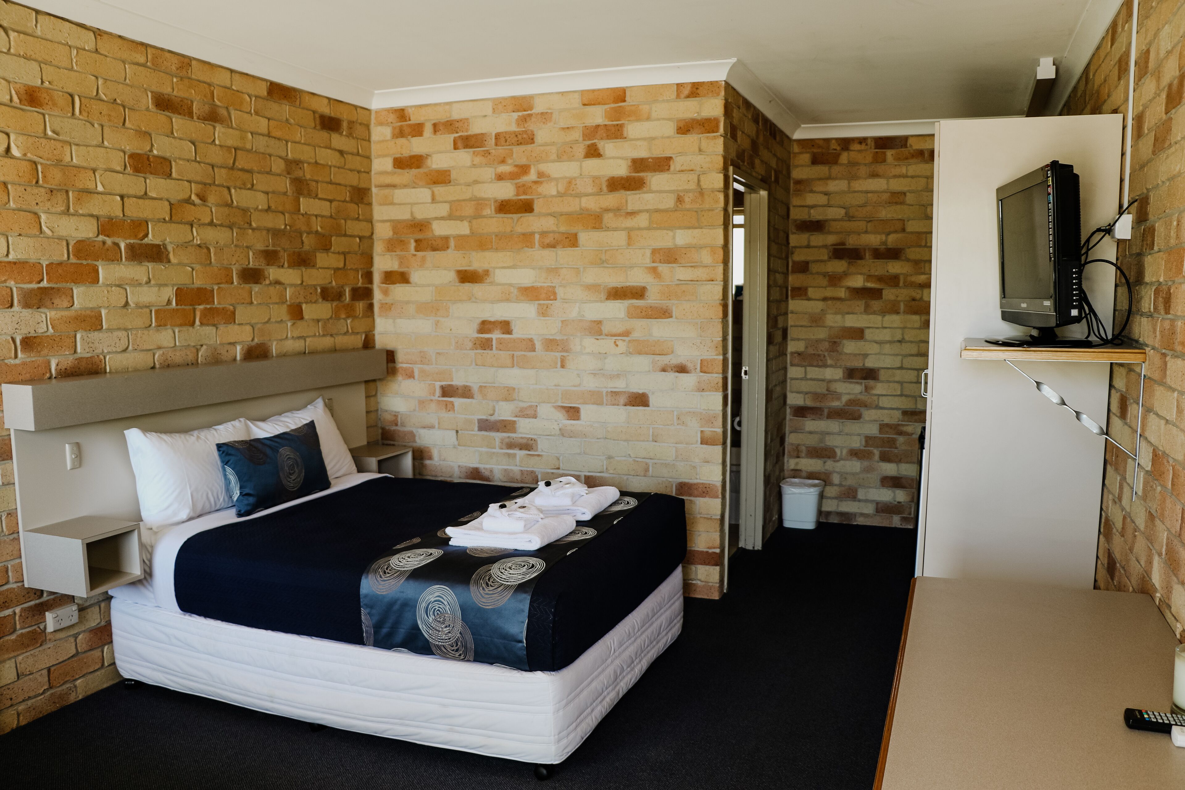 Quarto de hotel com parede de tijolos, cama com cabeceira, mesa de cabeceira, televisão sobre um suporte e porta para outro quarto.