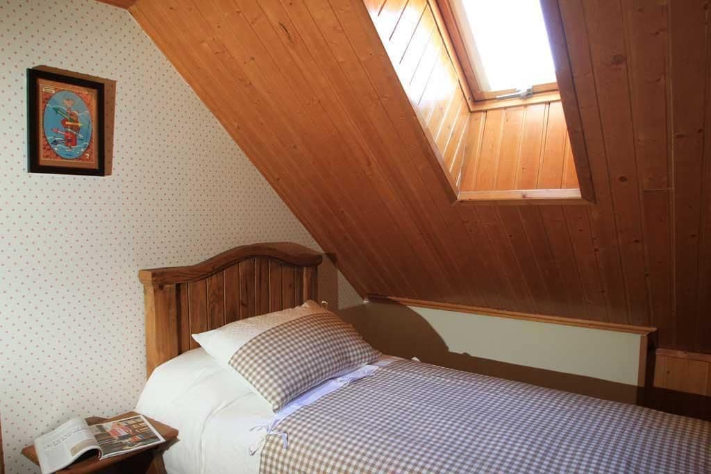 Quarto com teto inclinado, estrutura de cama de madeira e janela que permite entrada de luz natural.