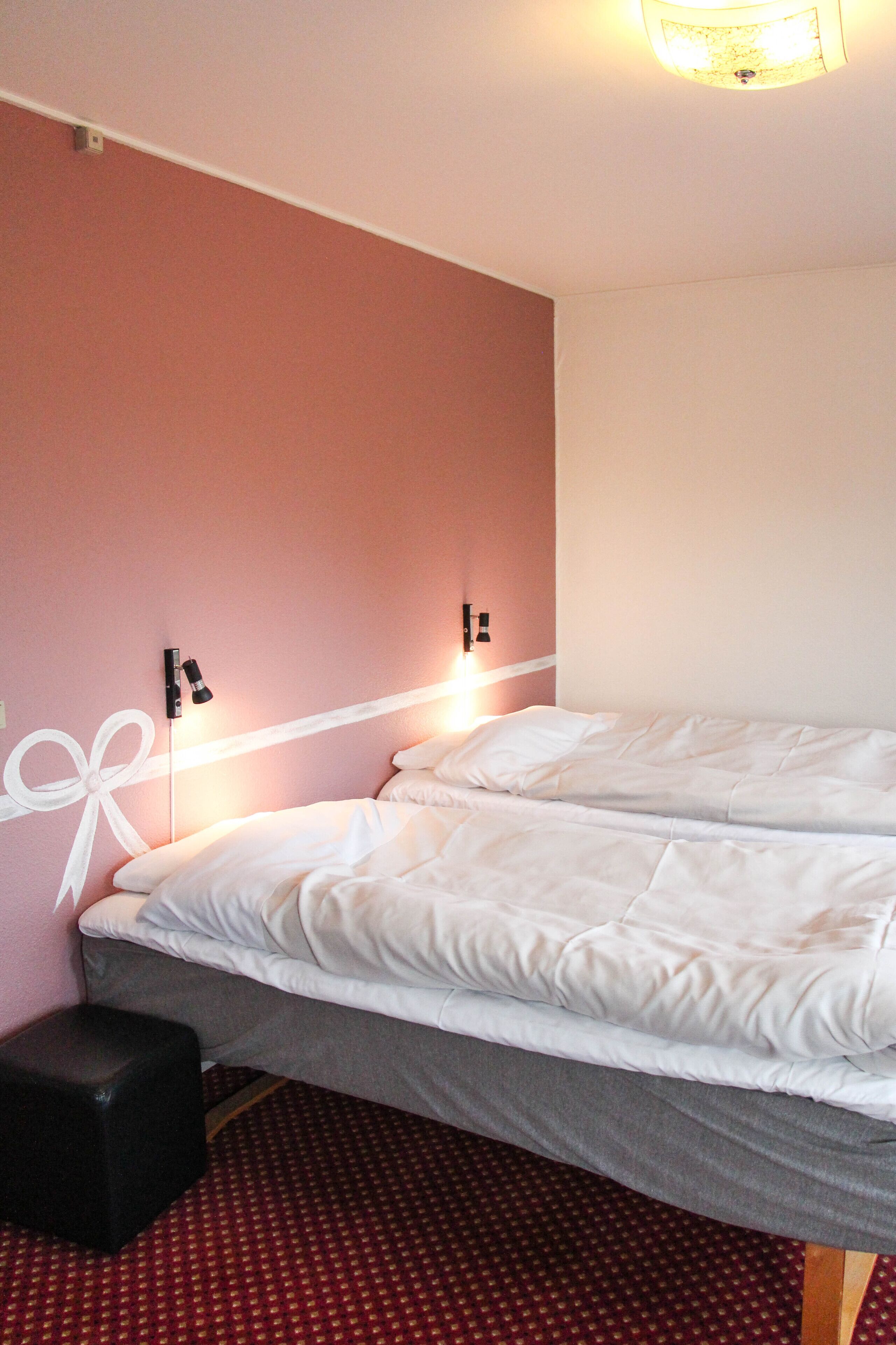 Cama de casal com roupa de cama branca, um banquinho preto pequeno e um painel decorado com uma fita branca.