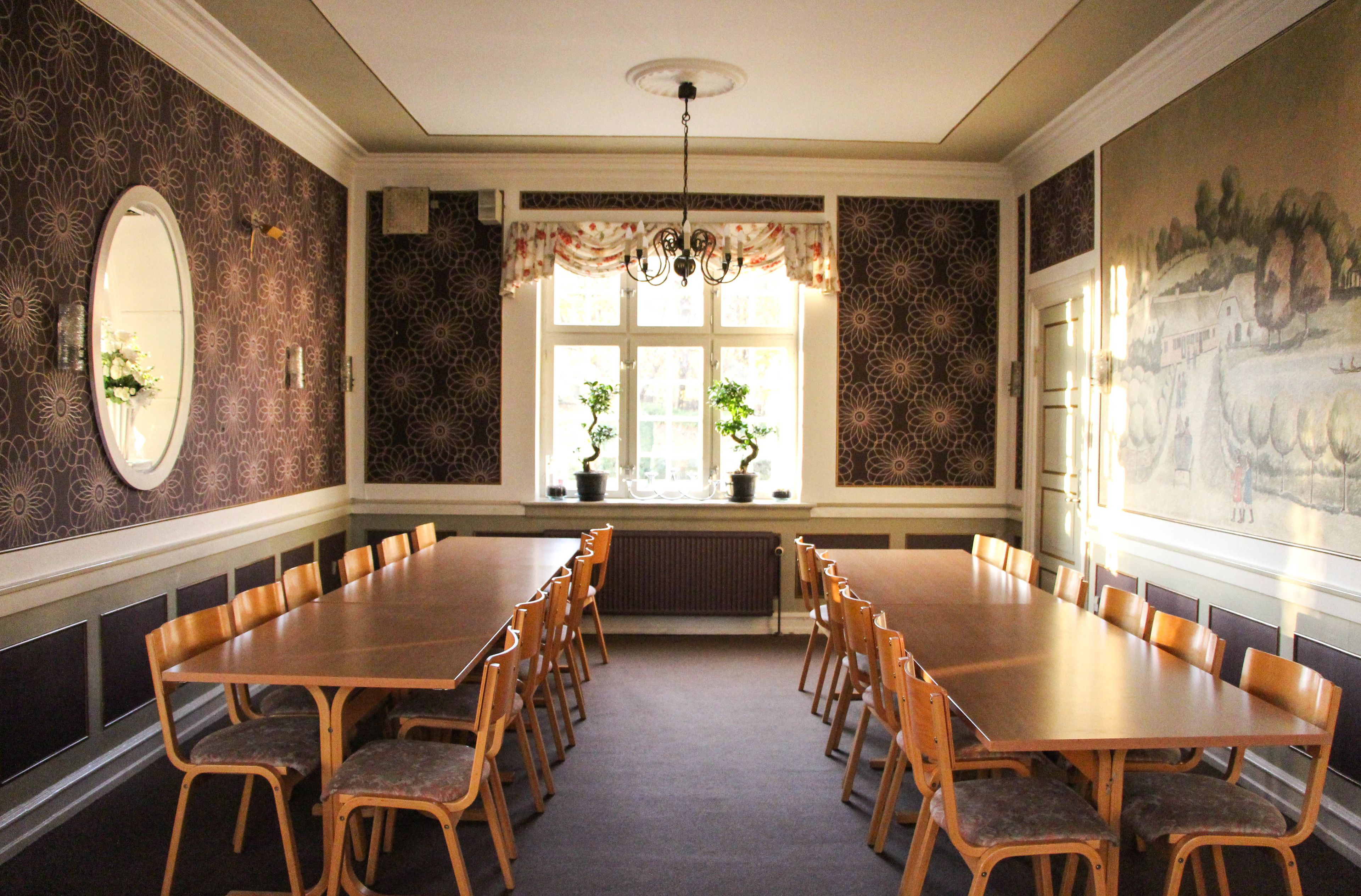 Uma sala de conferências com uma mesa grande e cadeiras, papel de parede estampado, um grande janelão com plantas e um candelabro.