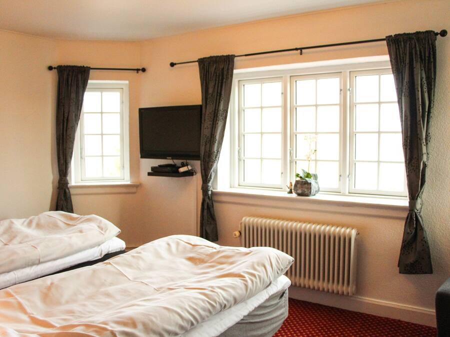 Quarto de hotel com duas camas, televisão na parede, radiador e janela com cortinas.