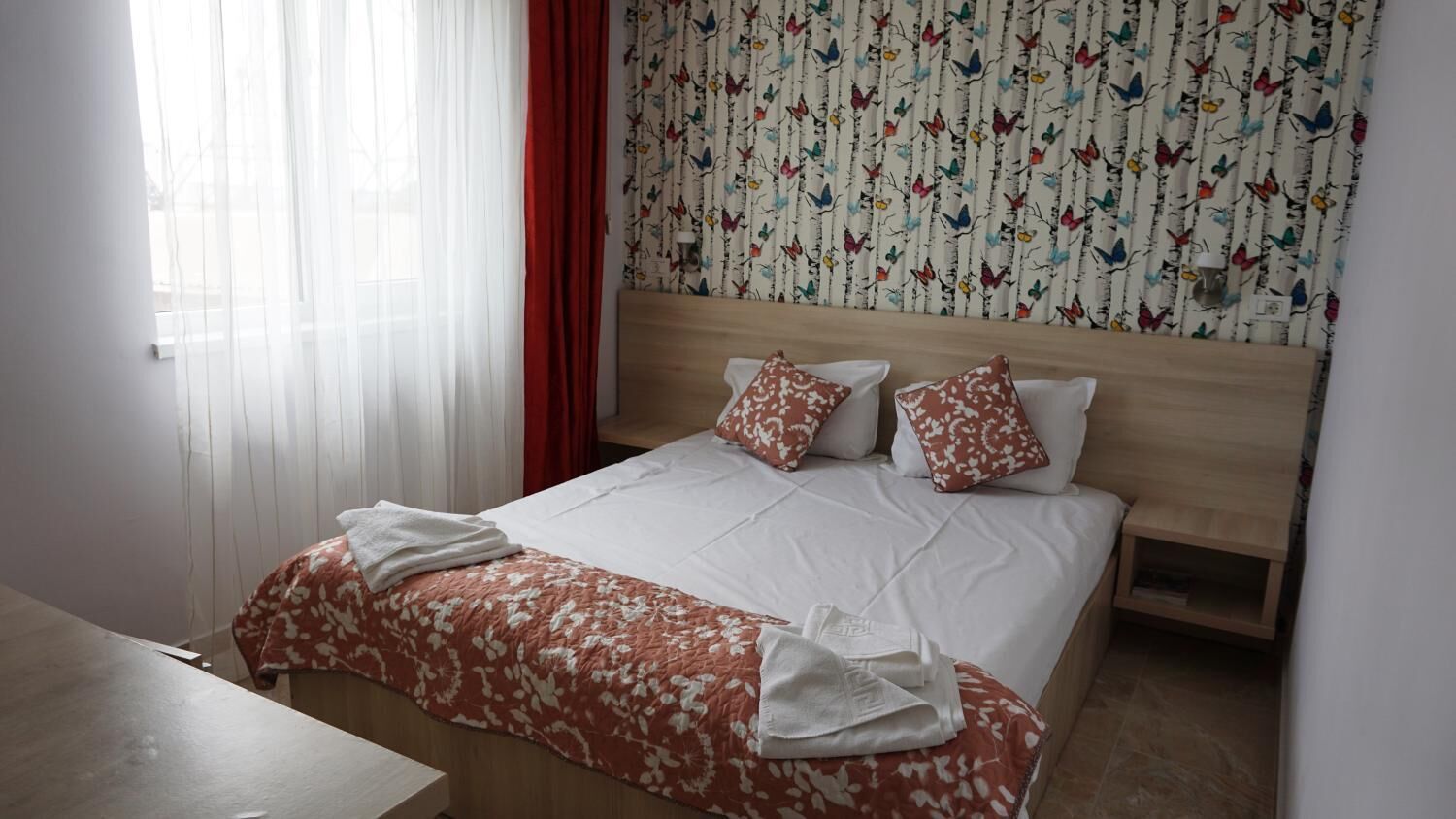 Cama bem arrumada com colcha floral e almofadas combinando, cabeceira de madeira e janela com cortinas leves.