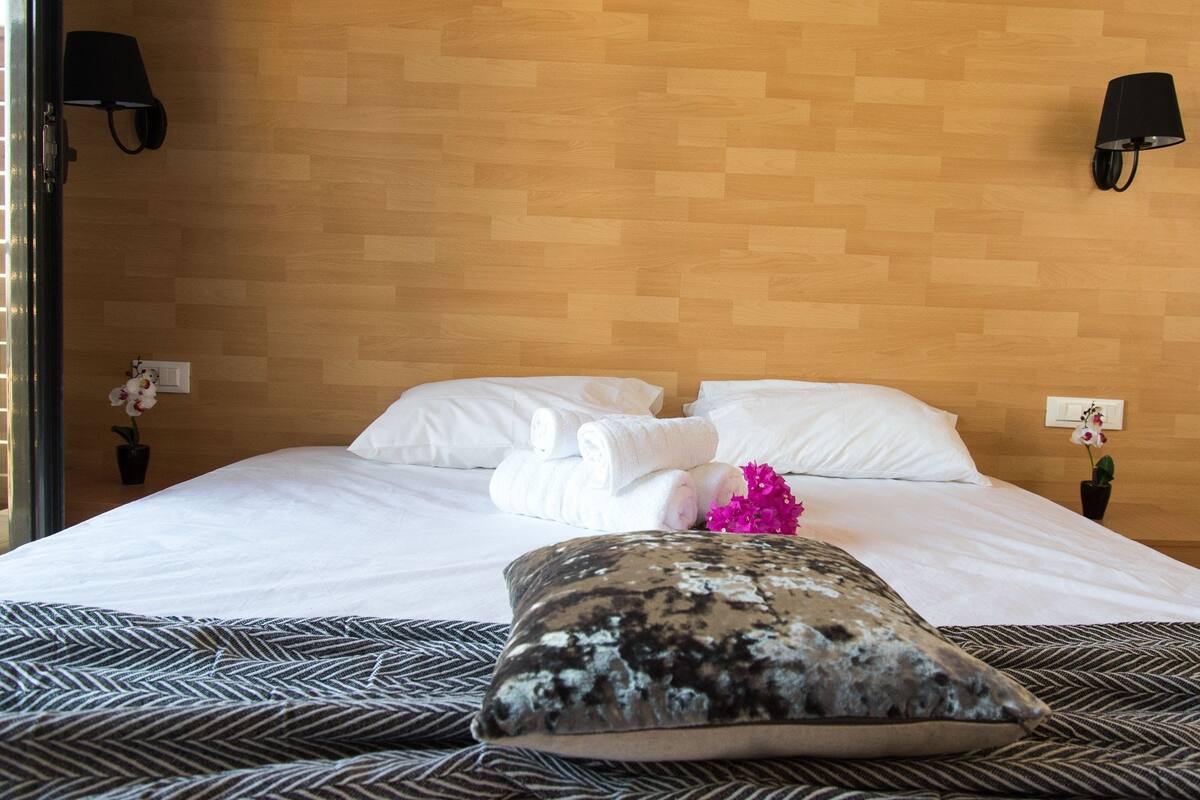 Quarto de hotel com cabeceira de madeira, roupa de cama branca, manta com padrão e duas orquídeas em vasos.