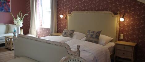 Quarto com cama grande, duas mesas de cabeceira, um criado-mudo, um banquinho e janela com cortinas.