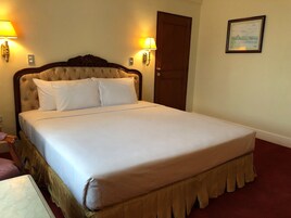 Uma cama bem arrumada com cabeceira, duas luminárias fixadas na parede, uma porta e um quadro emoldurado.
