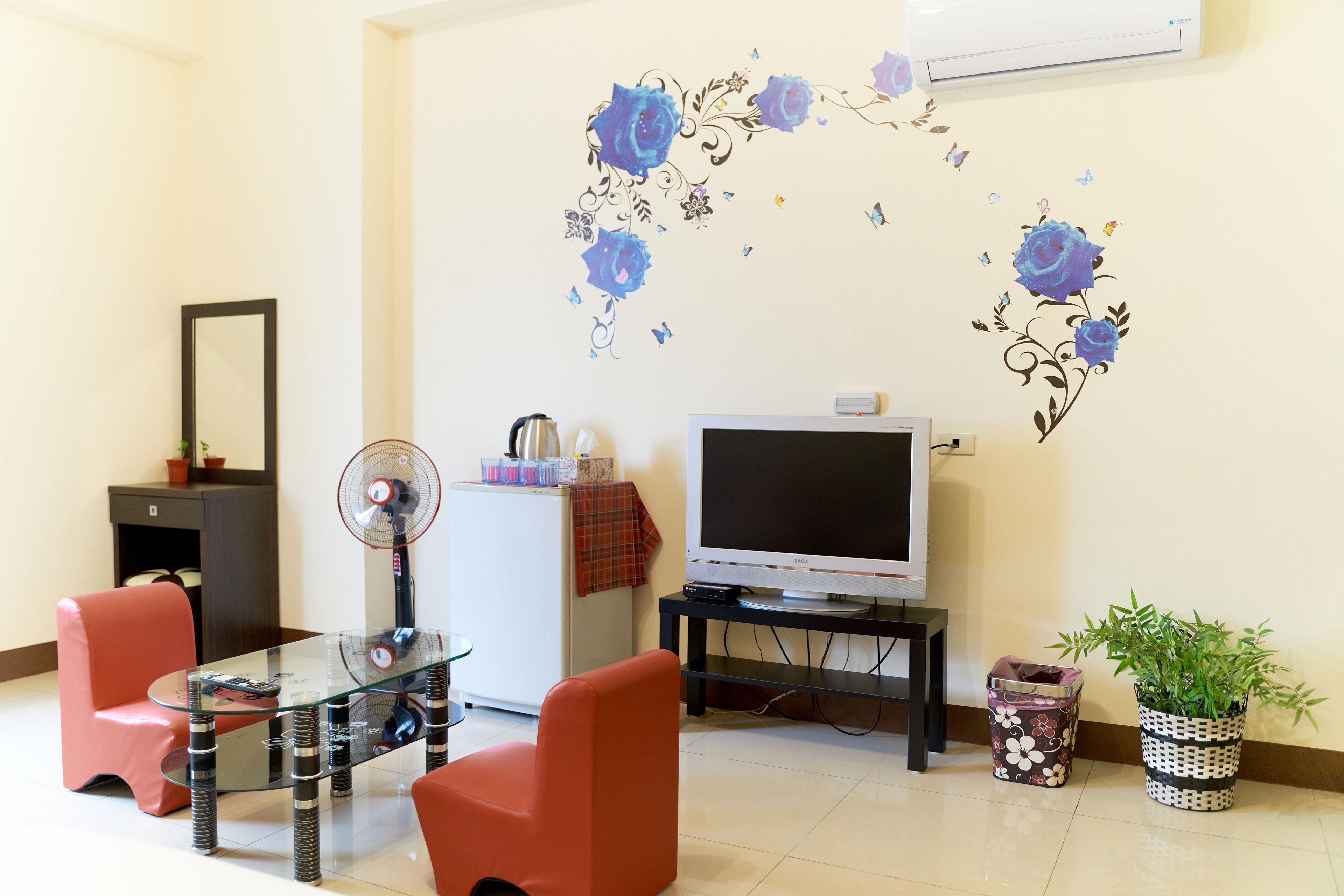 Sala de estar com mesa de vidro, cadeiras vermelhas, televisão, ventilador, frigorífico e autocolantes decorativos na parede.