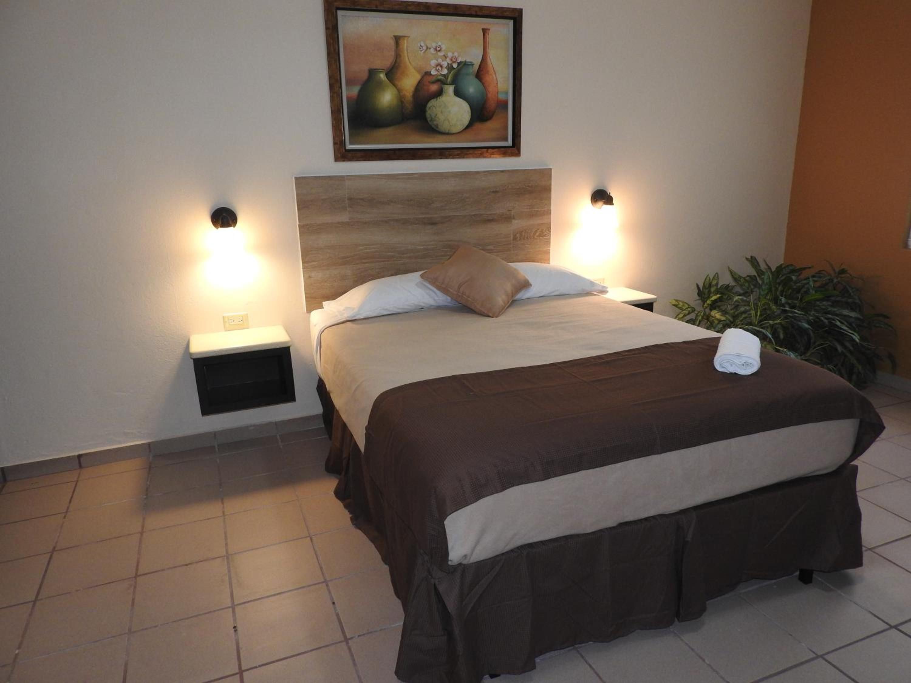 Cama bem arrumada com colcha marrom e um único travesseiro, com duas mesas de cabeceira com luminárias, cabeceira de madeira e um quadro acima da cama.