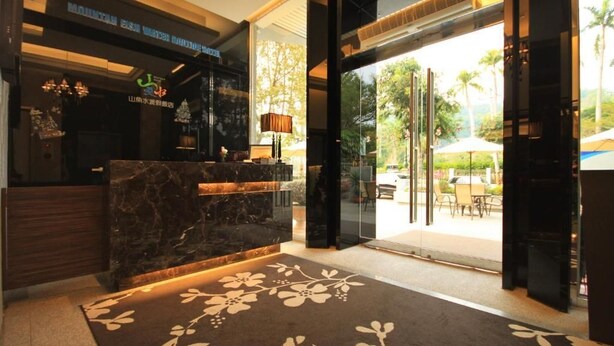 Uma área de recepção de hotel com balcão de mármore, carpete floral e vista para a área de estar externa através de portas de vidro.