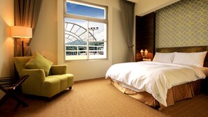 Quarto de hotel com cama, uma cadeira, abajur, janela com vista para uma ponte e papel de parede estampado.