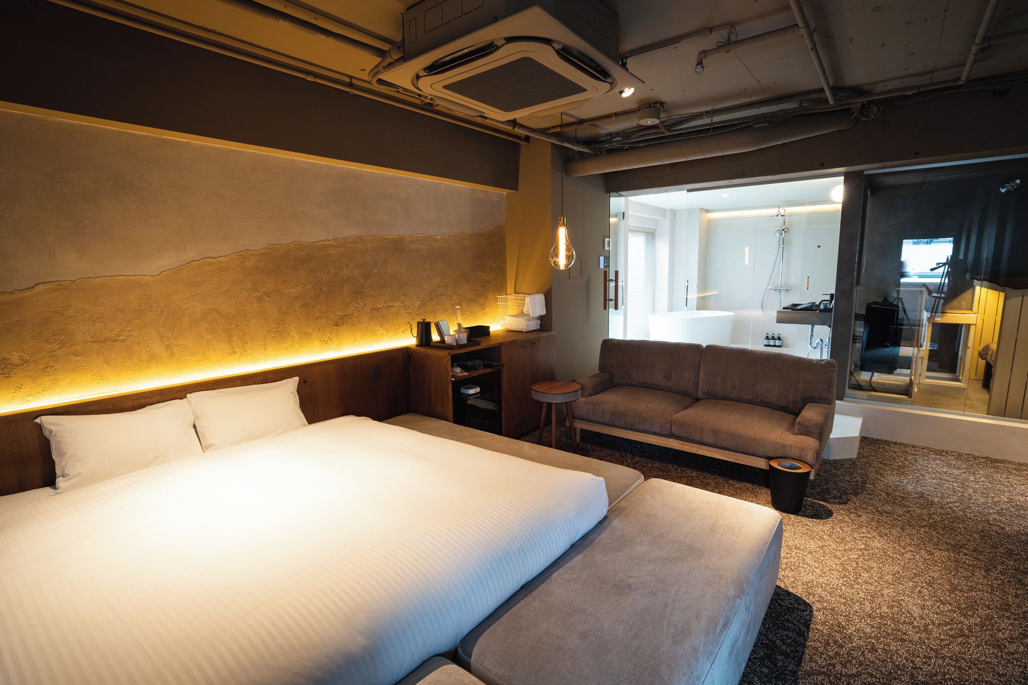 Quarto de hotel com uma cama grande, um sofá, uma mesa pequena e um banheiro visível por uma porta de vidro.