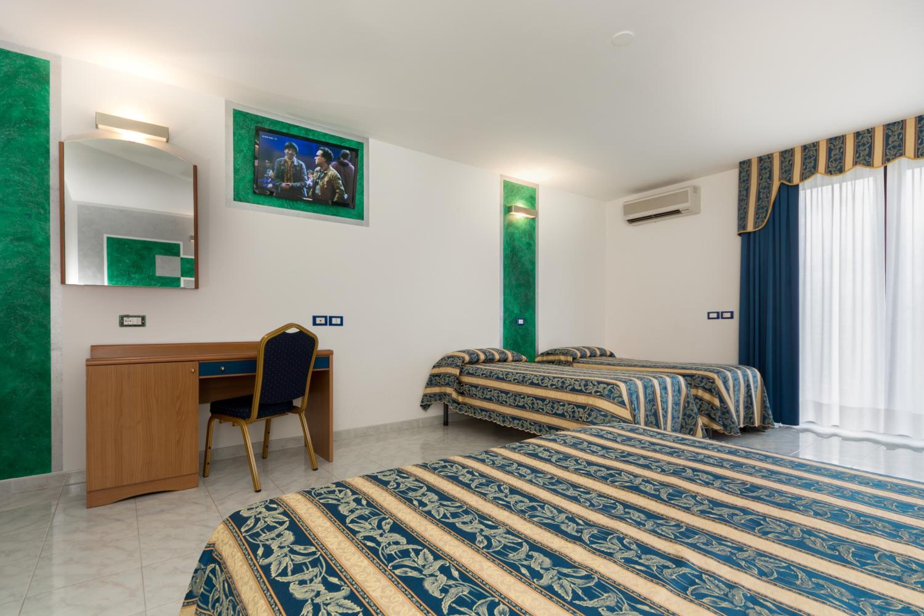 Quarto de hotel com escrivaninha, duas camas, espelho, quadro emoldurado e janela com cortinas.