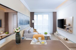 Quarto de hotel moderno com sofá, mesa de jantar com um vaso de flores, televisão e uma grande janela com cortinas.