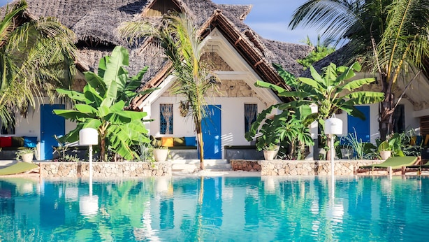 Vista da piscina de um resort, com um edifício de telhado de palha, palmeiras e uma piscina azul cristalina.