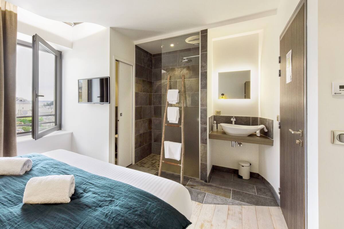 Quarto de hotel com cama, banheiro com chuveiro e vista pela janela.