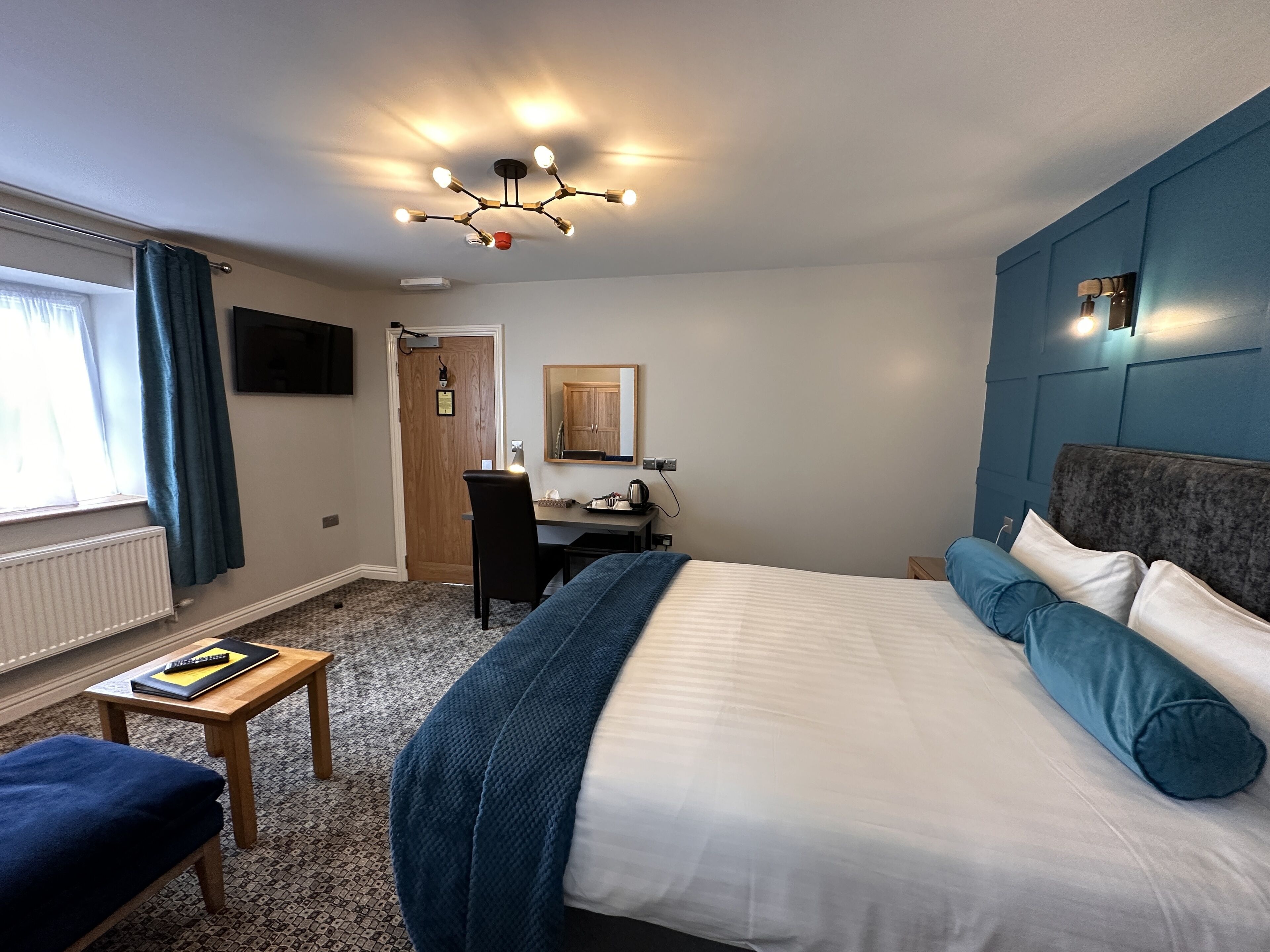 Quarto de hotel com cama, escrivaninha, cadeira, mesa pequena e luminária no teto.