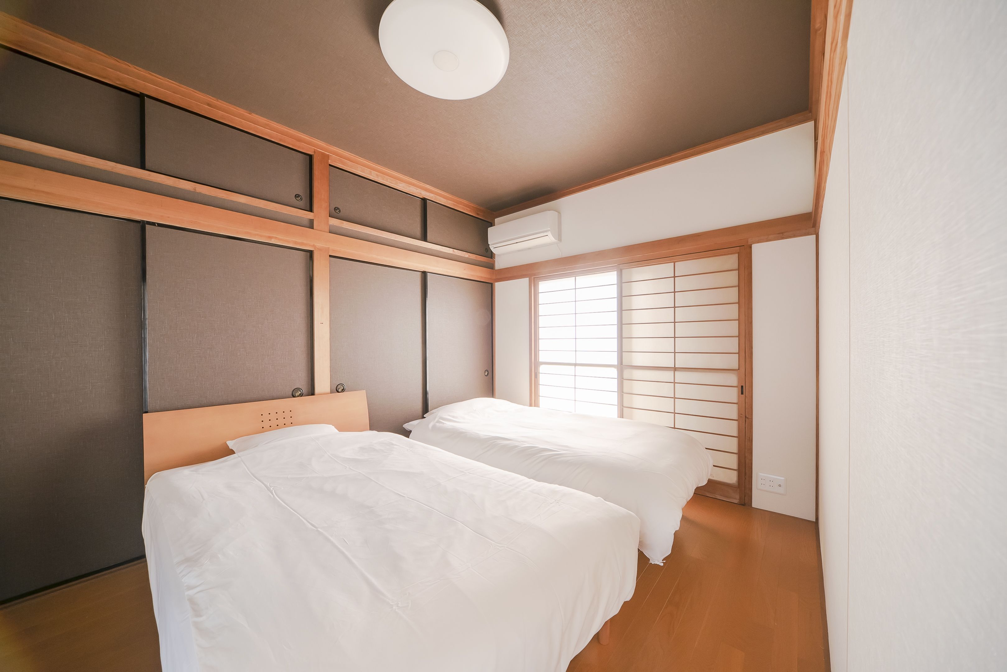 Quarto com duas camas, piso de madeira, luminária no teto e janela com persianas.