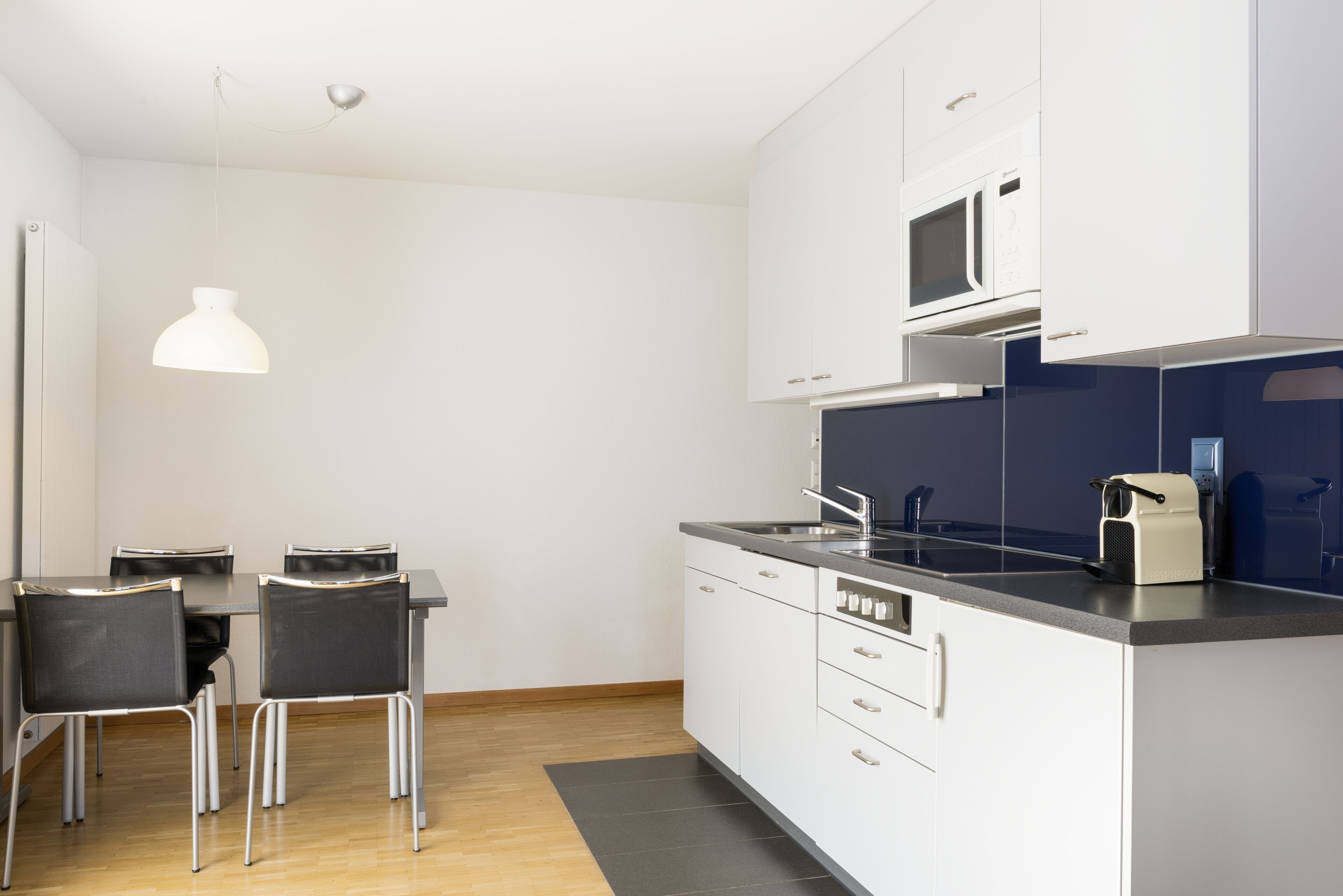 Cozinha moderna com armários brancos, bancada preta e micro-ondas. Duas cadeiras estão dispostas em torno de uma mesa, e uma luminária pendente ilumina o teto.