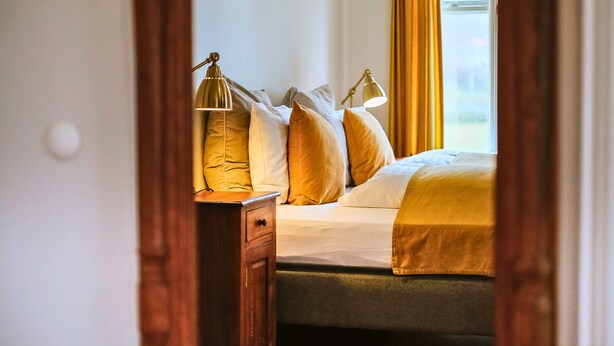 Cama bem arrumada com colcha verde e várias almofadas, mesa de cabeceira de madeira com duas luminárias, e janela com cortinas amarelas.