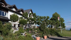 Uma casa de dois andares com telhado inclinado, janelas amplas e um jardim bem cuidado com diversas plantas e árvores em vasos.