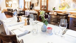 Uma área de refeições com mesas preparadas para dois, com toalhas brancas, copos de vinho transparentes e um vaso com flores.