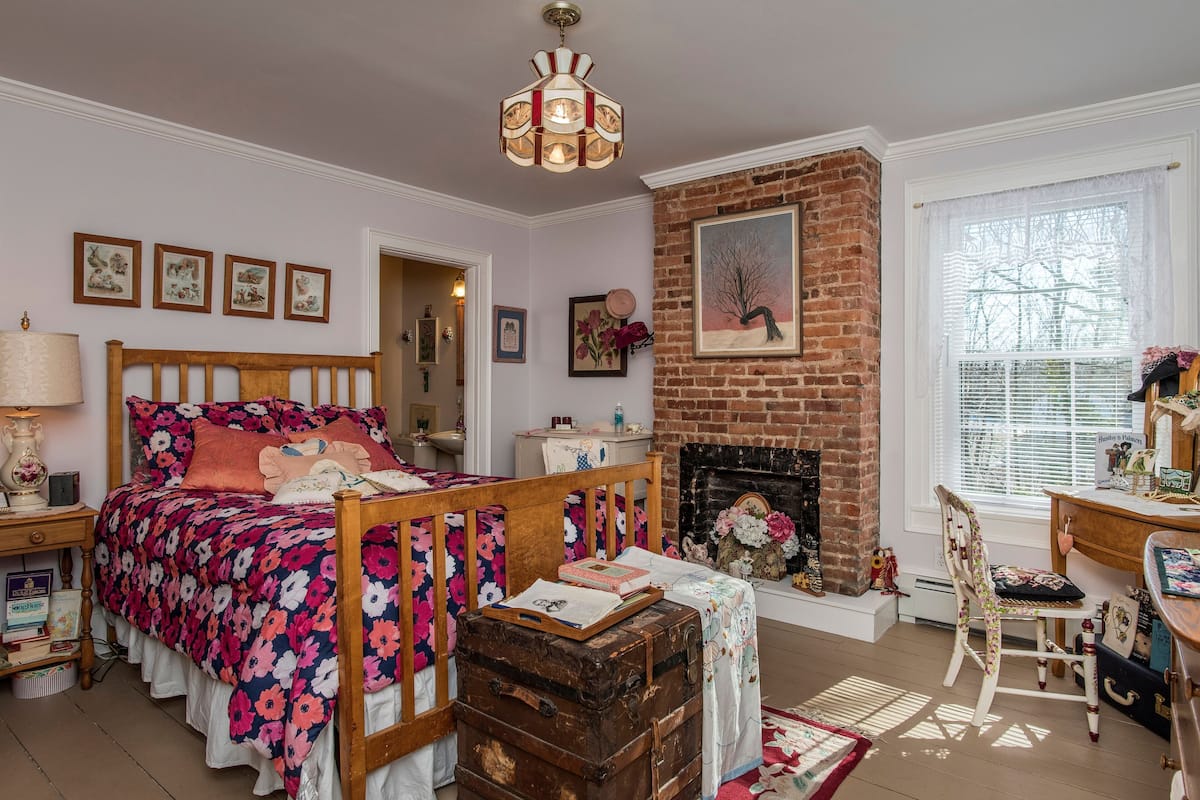 Quarto aconchegante com cama de madeira, lareira de tijolos e uma janela grande com persianas.