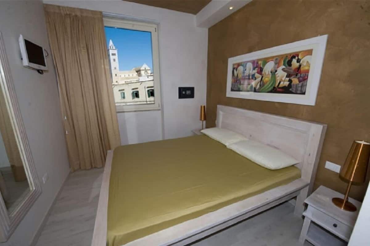 Um quarto com cama, janela com cortinas, um quadro na parede e mesa de cabeceira com abajur.
