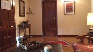 Quarto com porta de madeira, um quadro pendurado na parede, uma mesa pequena com uma caneca, um cesto de flores secas e um tapete vermelho.