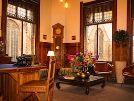 Quarto com escrivaninha de madeira, relógio, sofá, mesa de centro e vista de um edifício por grandes janelas.