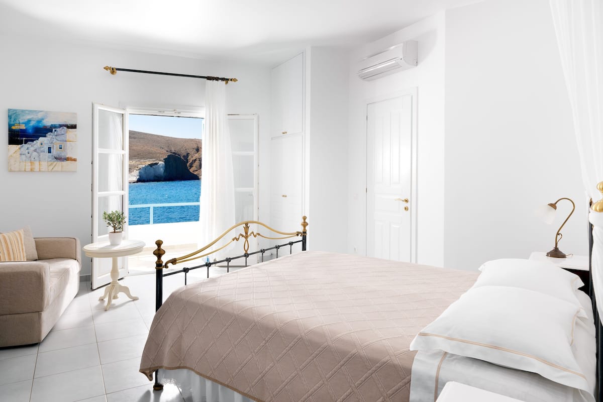 Quarto com cama, mesa de cabeceira, cadeira, janela com vista para o mar e um quadro na parede.