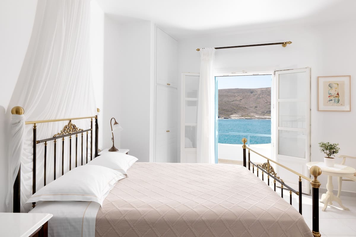 Um quarto com uma cama, uma mesa de cabeceira, uma luminária, uma janela com vista para o mar e uma porta que dá para outro cômodo.