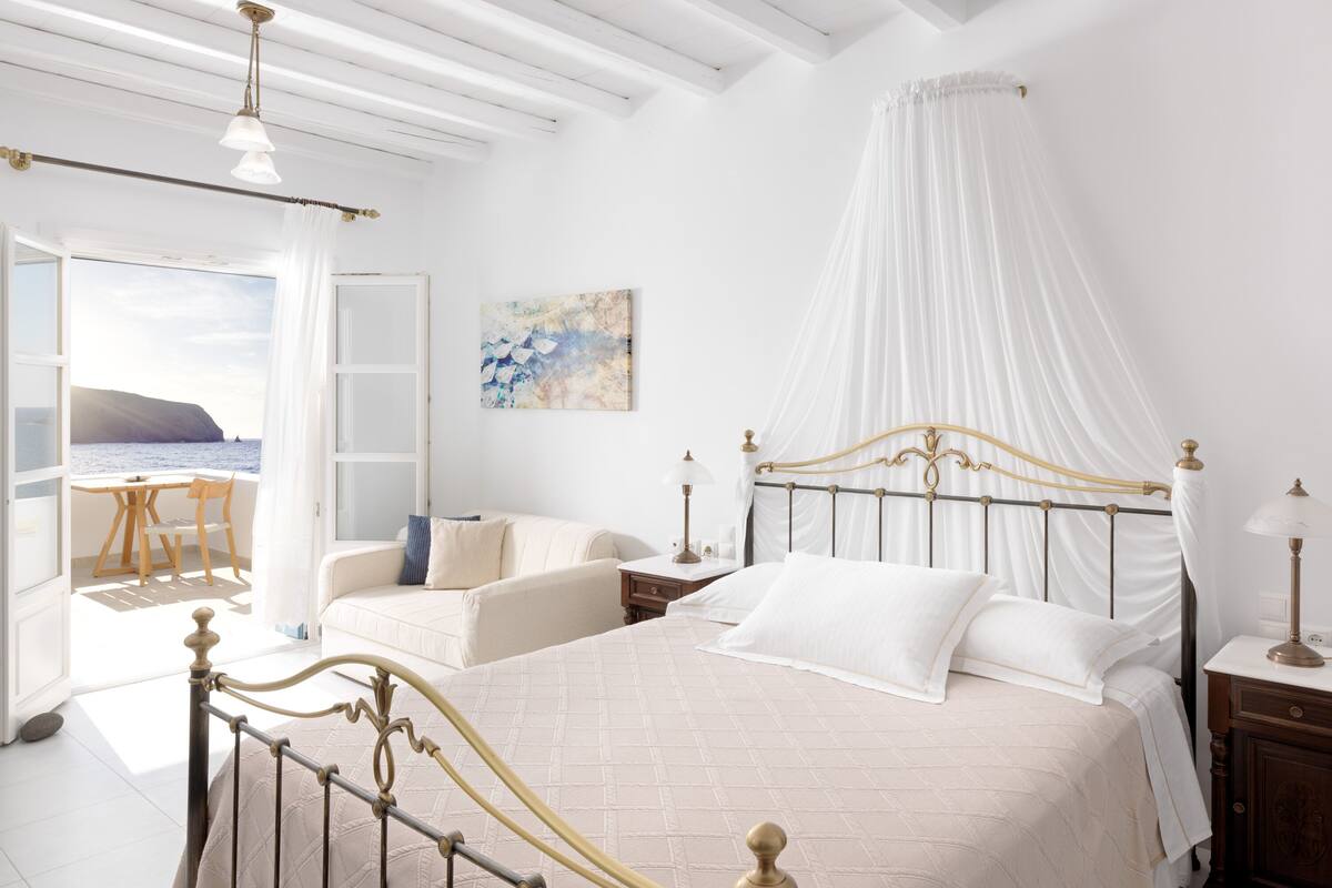 Quarto com cama, mesas de cabeceira, uma cadeira e vista para o mar.