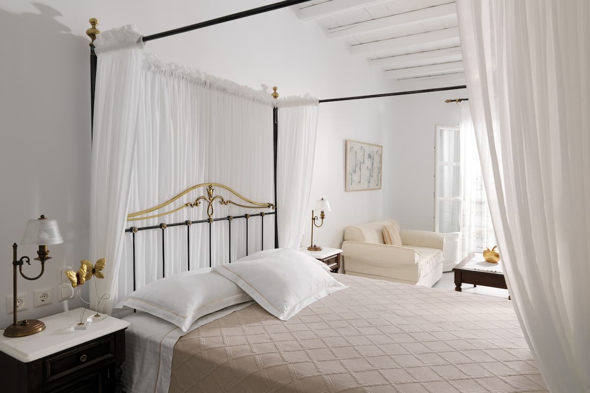 Uma cama de dossel com lençóis brancos, uma mesa de cabeceira com abajur e um sofá cor de creme em um quarto claro com paredes brancas e teto com vigas aparentes.